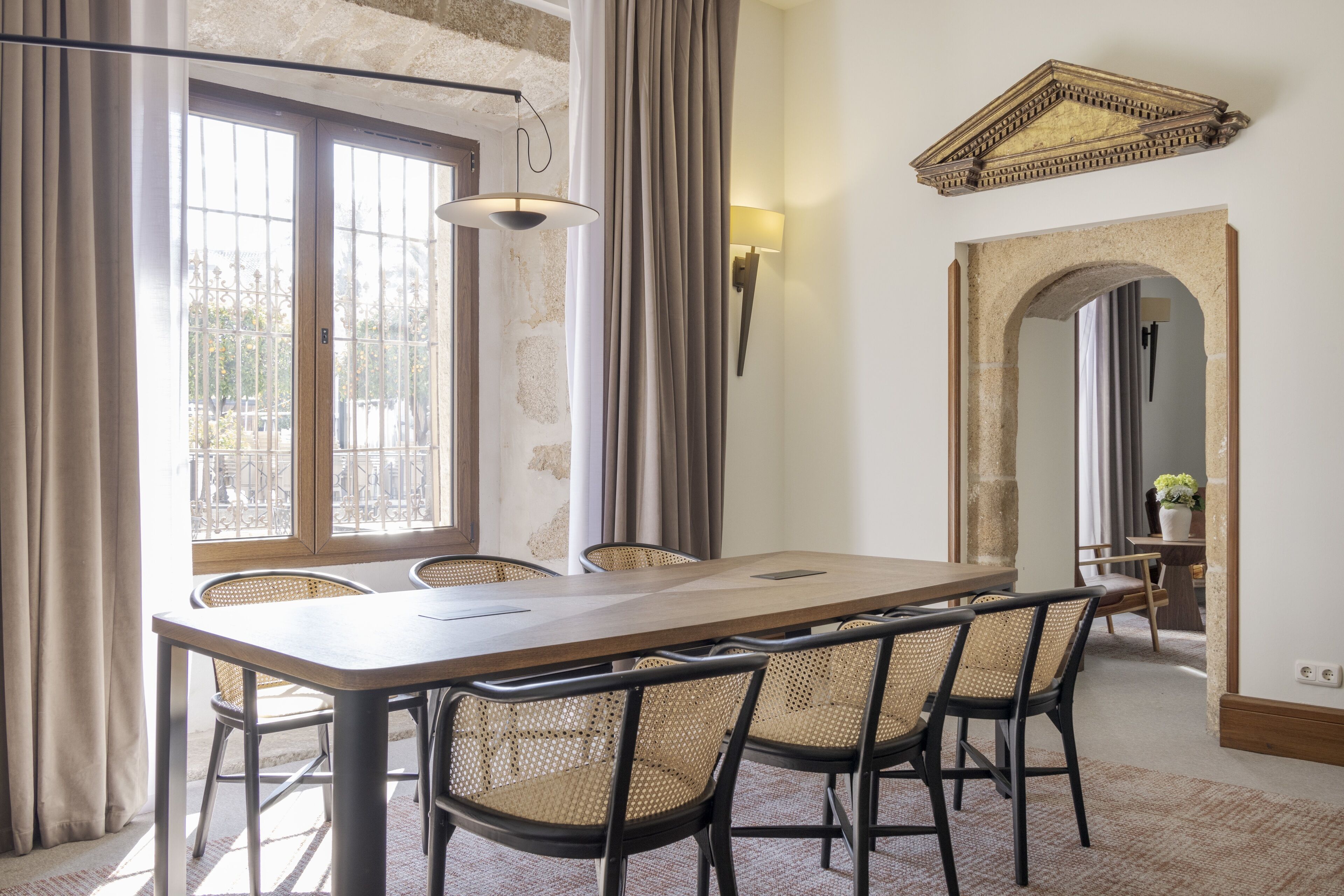 Uma área de refeições com mesa de madeira e cadeiras de vime, uma janela grande com cortinas e um aplique de parede.