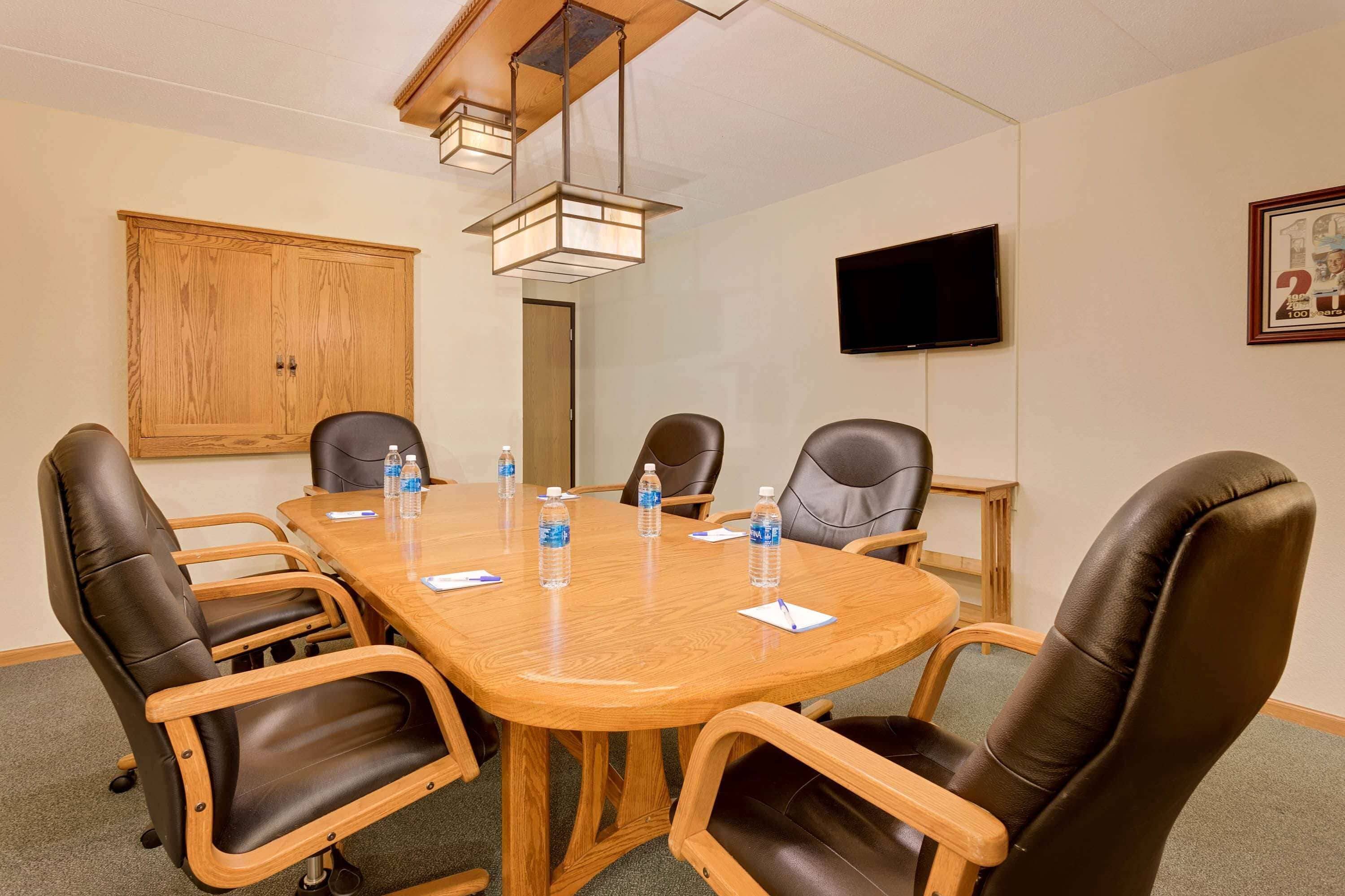 Uma sala de conferências com mesa de madeira, cadeiras de couro preto, garrafas de água, uma televisão e um armário de madeira.