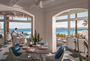 Restaurante com vista para o mar, com mesas com almofadas azuis, um garçom e um ventilador de teto.