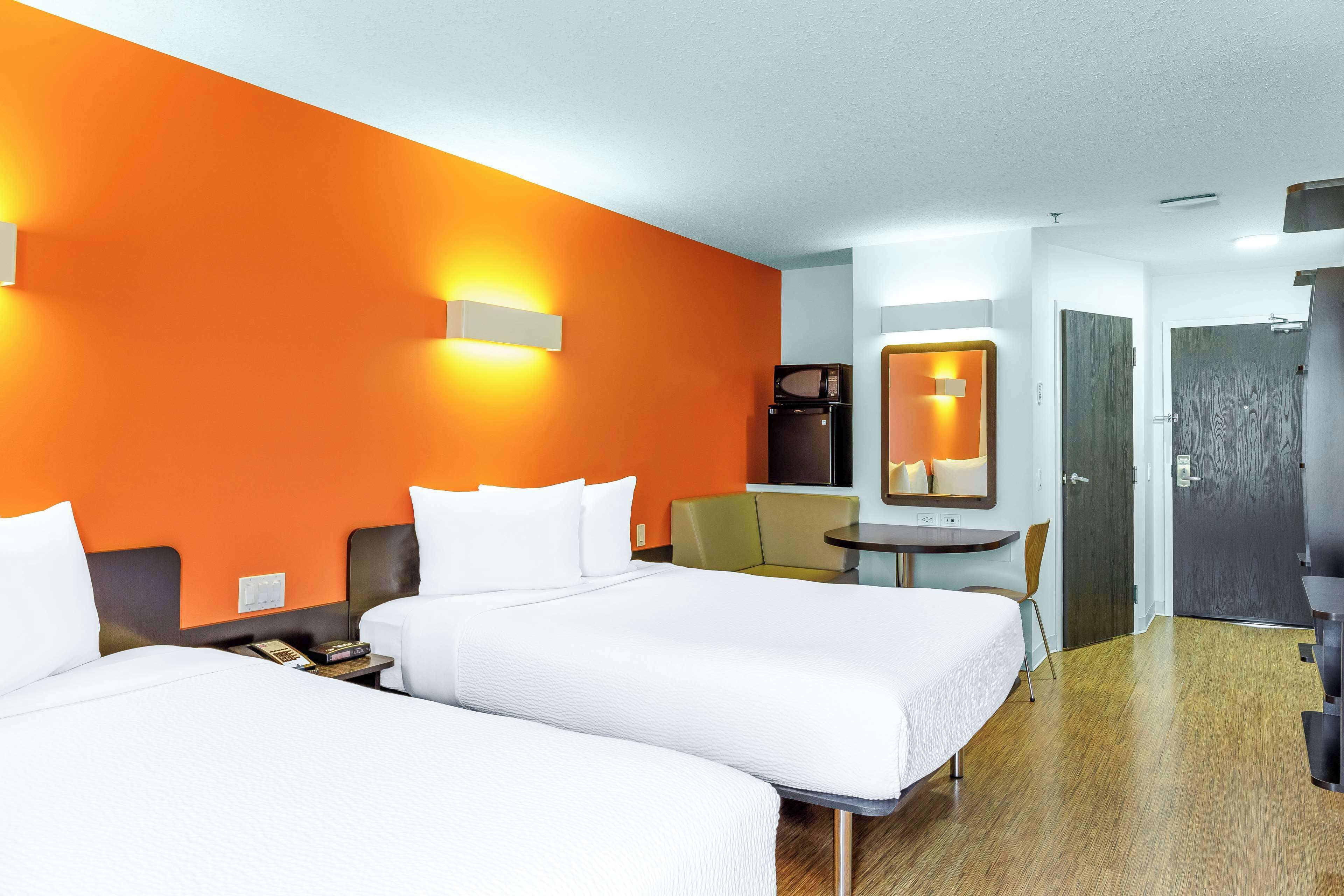 Quarto de hotel com duas camas, uma mesinha, uma cadeira, micro-ondas e uma porta.