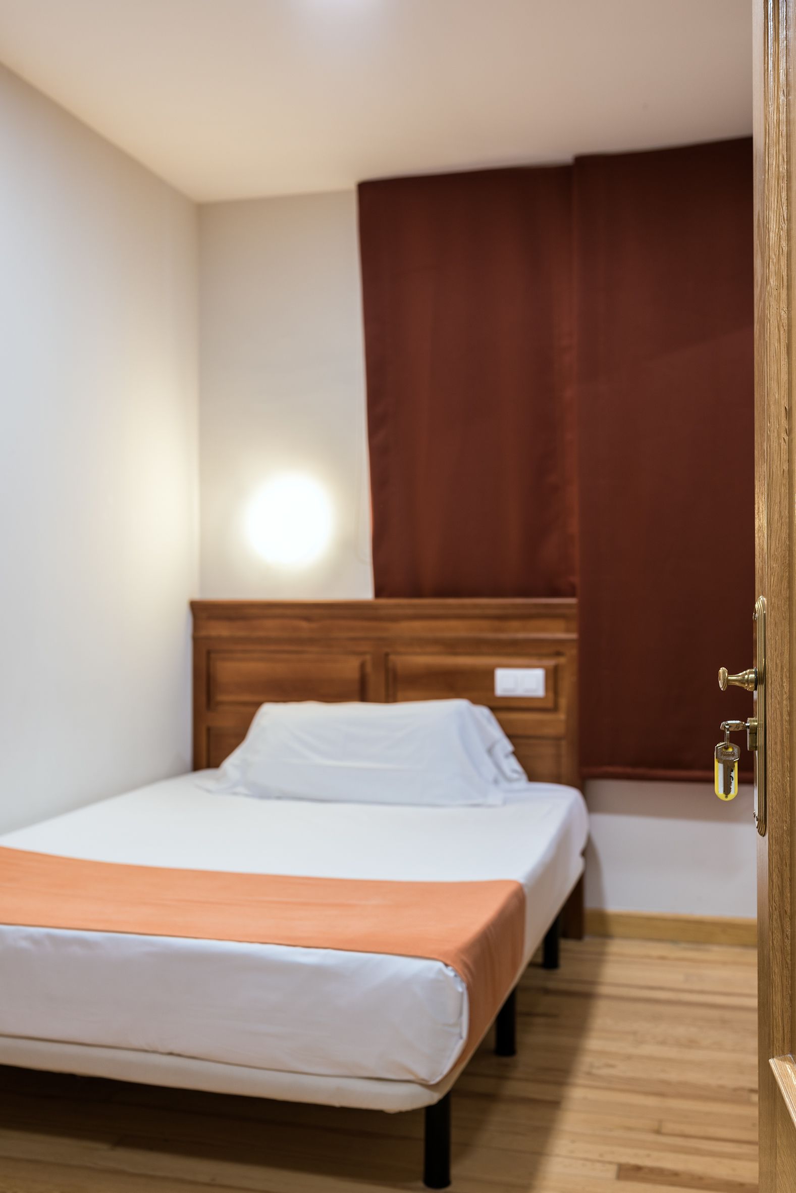 Quarto de hotel com cama de madeira, roupa de cama branca, cabeceira de madeira, cortina marrom e chão de madeira.