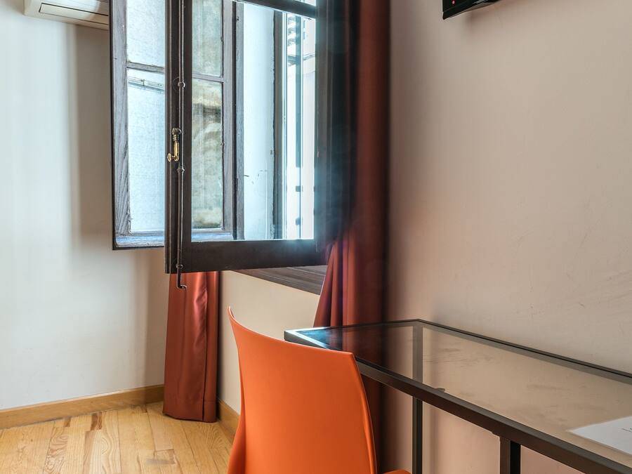 Quarto com piso de madeira, mesa de vidro, cadeira laranja, TV na parede e janela com cortinas.