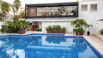 Um edifício moderno com fachada de vidro, piscina e palmeiras.