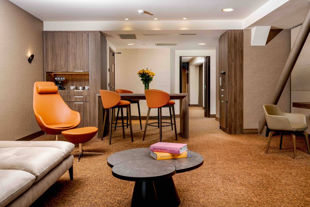 Quarto de hotel com sofá, mesa de jantar com cadeiras, mesinha redonda com livros e cozinha compacta.