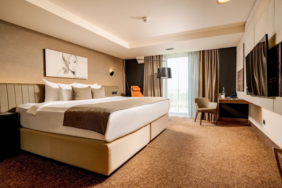 Quarto de hotel com cama grande, escrivaninha, cadeira, televisão e janela com cortinas.