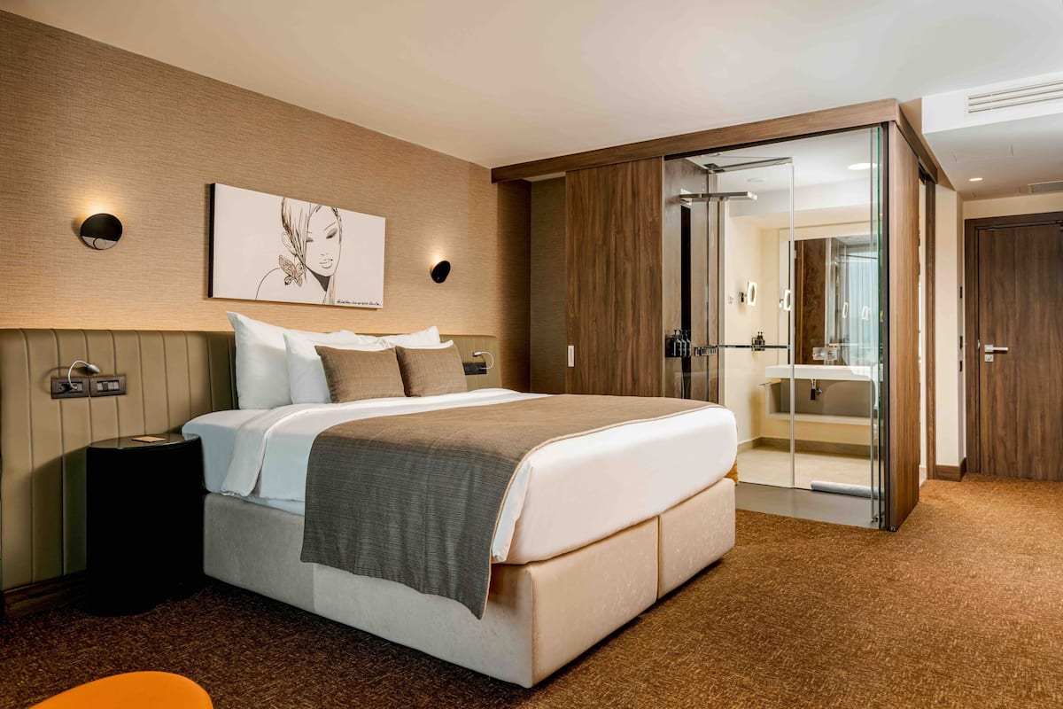 Quarto de hotel moderno com uma cama grande, mesa de cabeceira, armário de madeira e chuveiro envidraçado.