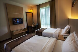 Quarto de hotel com duas camas, uma escrivaninha, televisão e uma janela grande com cortinas.