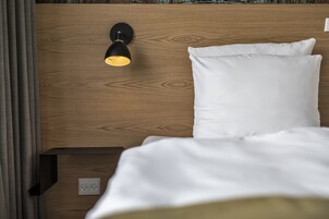 Uma cama bem arrumada com roupa de cama branca, cabeceira de madeira com luz fixada na parede e uma tomada elétrica abaixo.