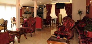 Um saguão de hotel com área de refeições, mobiliário estofado vermelho, uma TV de tela plana e um lustre.