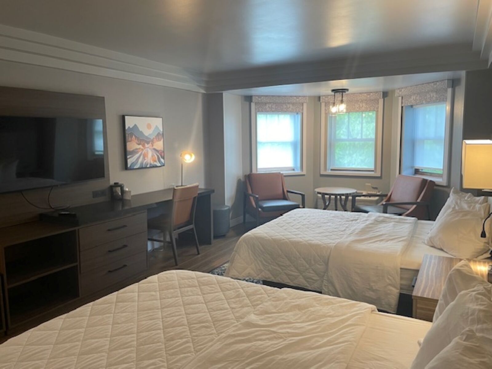 Quarto de hotel com duas camas, uma escrivaninha com cadeira, televisão e um quadro na parede.