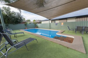 Piscina com área coberta por sombra, rodeada por grama sintética e um rodapé de tijolos.