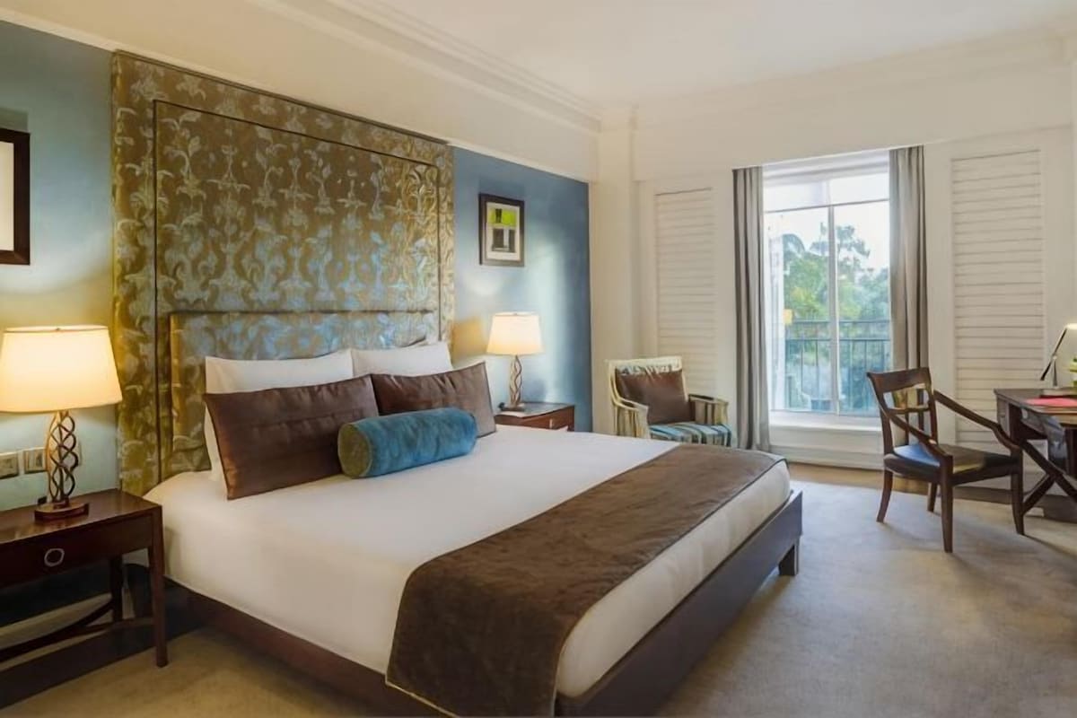 Quarto de hotel com uma cama grande, duas mesas de cabeceira com abajures, uma cadeira, uma escrivaninha e uma janela com vista.