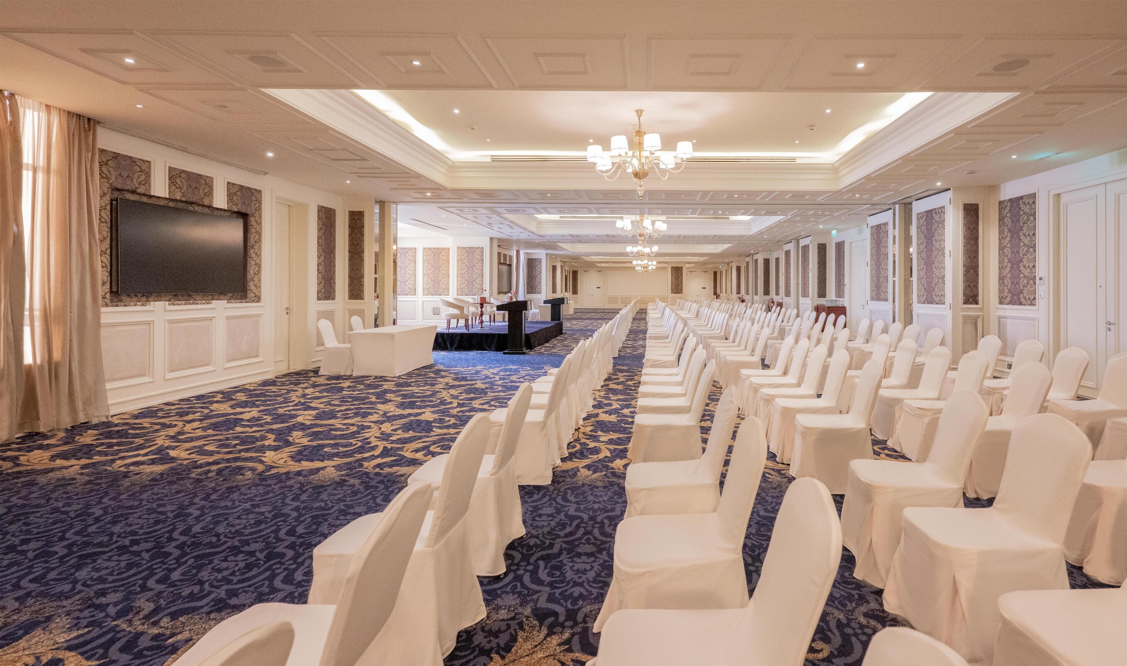 Uma sala de conferência com fileiras de cadeiras brancas, um palco com um pódio e uma tela grande.