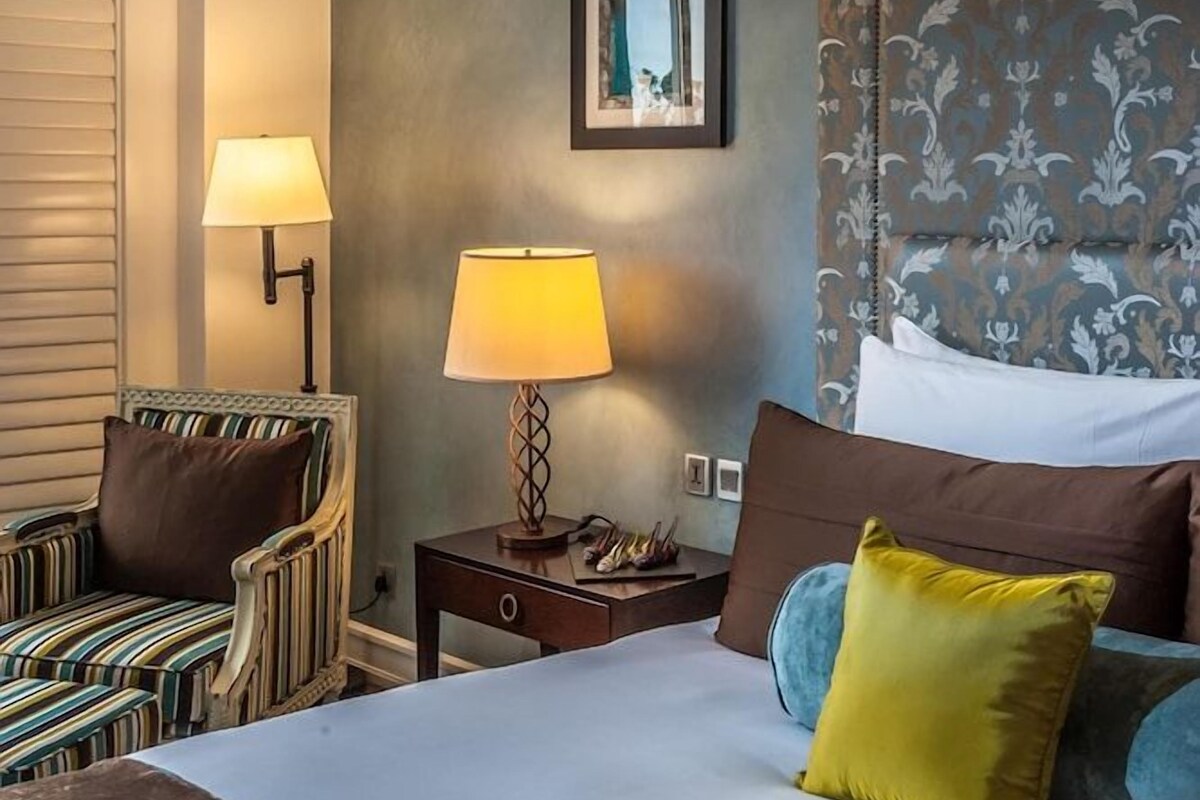 Quarto de hotel com cama, duas luminárias de cabeceira, uma poltrona, uma mesinha e um quadro na parede.