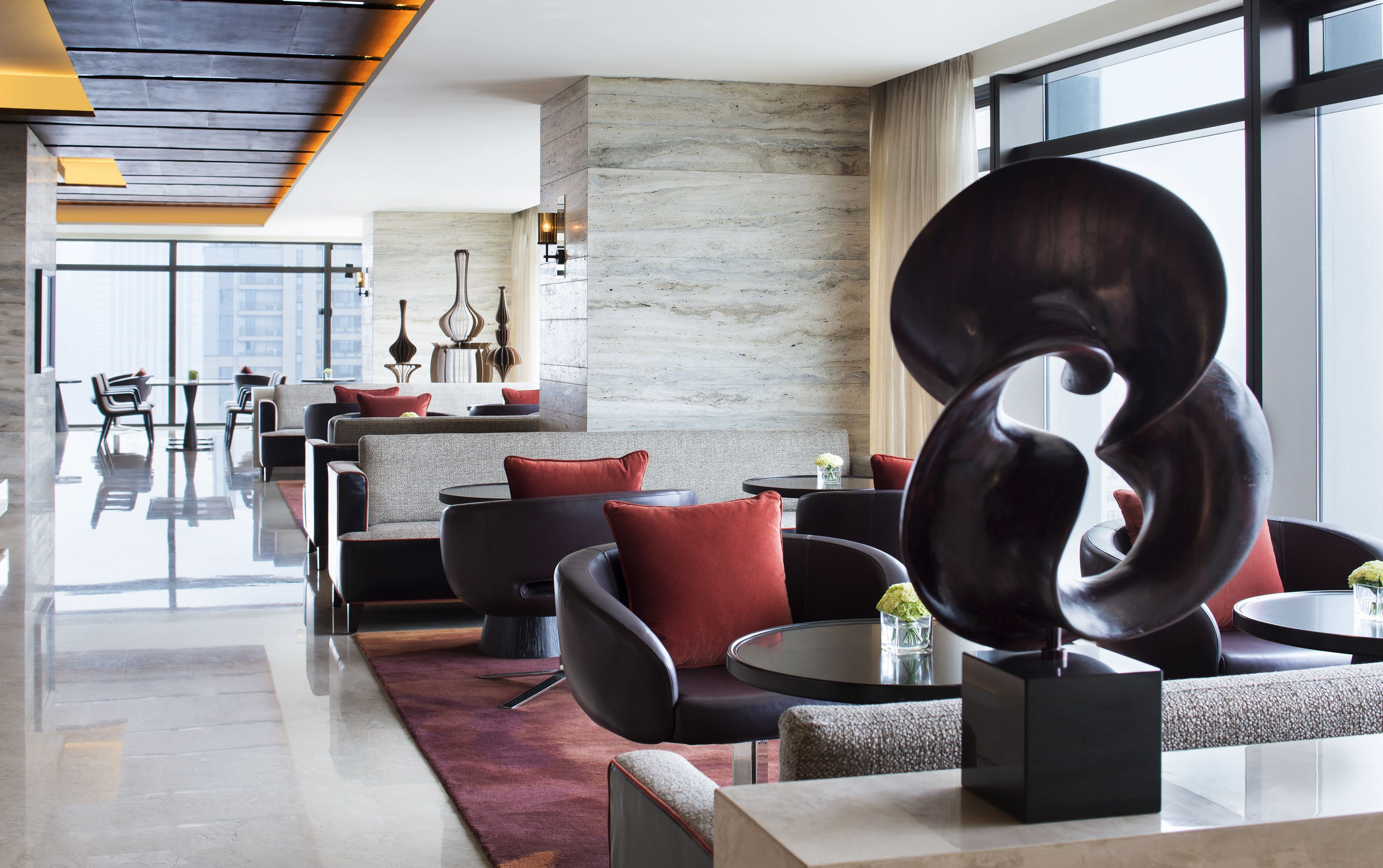 Um hall de hotel moderno com uma receção de mármore, áreas de estar com almofadas vermelhas e uma grande escultura abstrata.