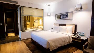 Quarto de hotel com uma cama grande, mesa de cabeceira com telefone e garrafas de água, uma cadeira, janela com persianas e um banheiro visível através da janela.
