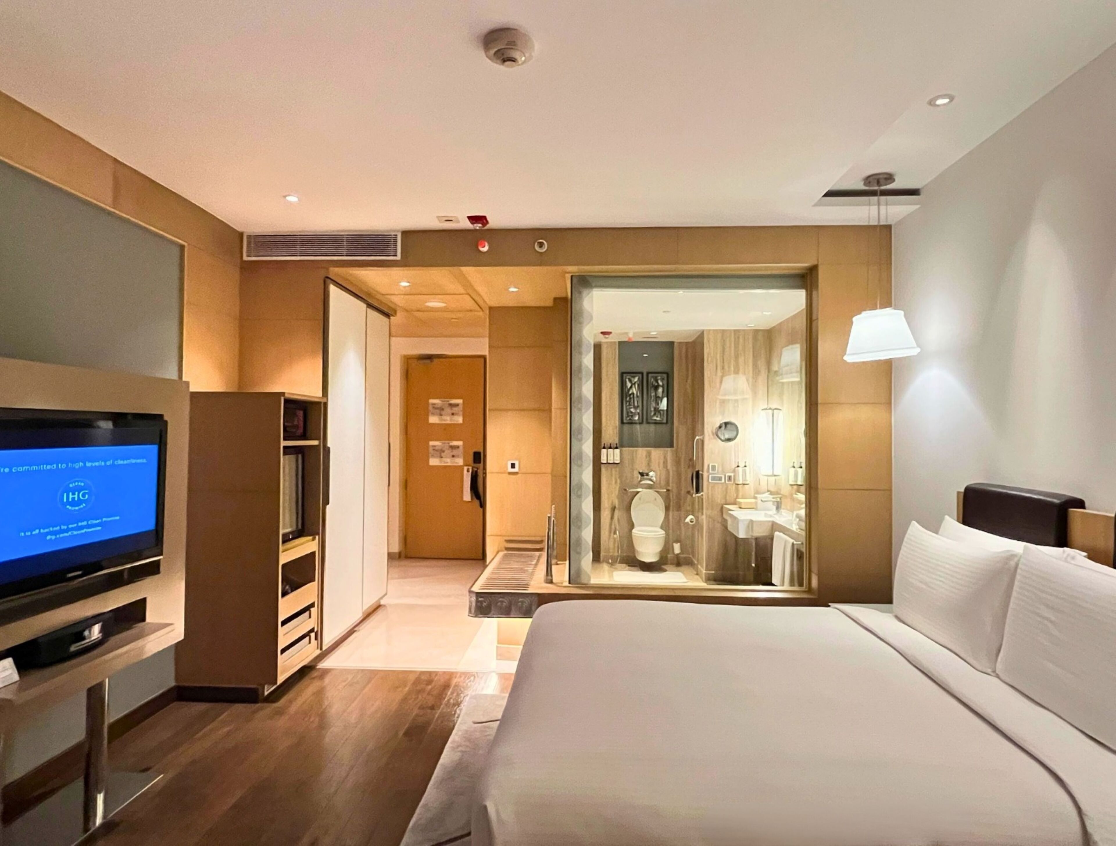 Quarto de hotel com cama, televisão, escrivaninha e banheiro visível através de uma porta aberta.