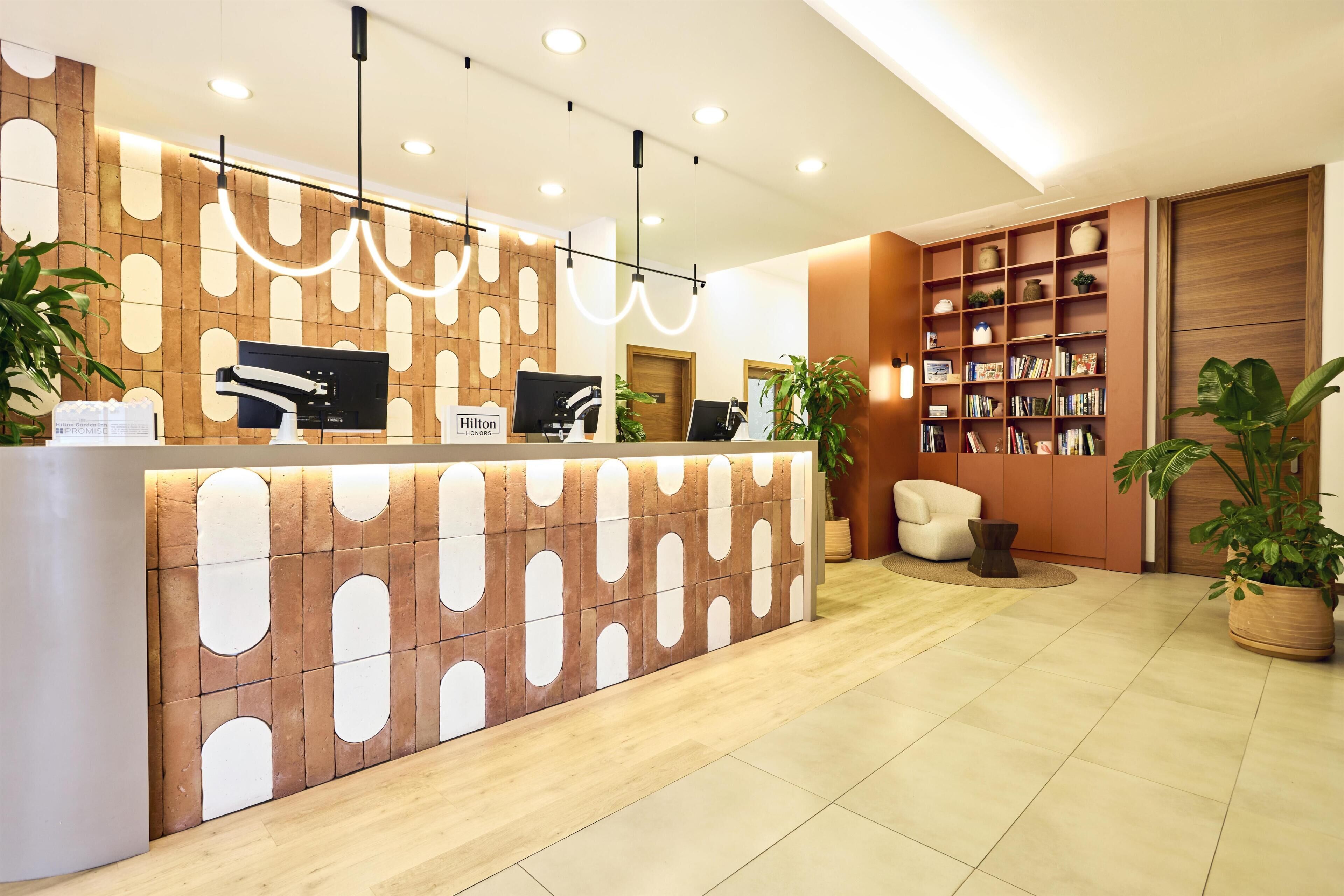 Uma área de recepção de hotel com um balcão de madeira, dois monitores de computador, uma área de estar com uma cadeira e uma estante cheia de livros.