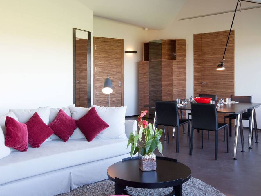 Uma sala de estar moderna com um sofá branco, almofadas vermelhas, uma mesa de centro preta e uma área de jantar com uma mesa e cadeiras de madeira.