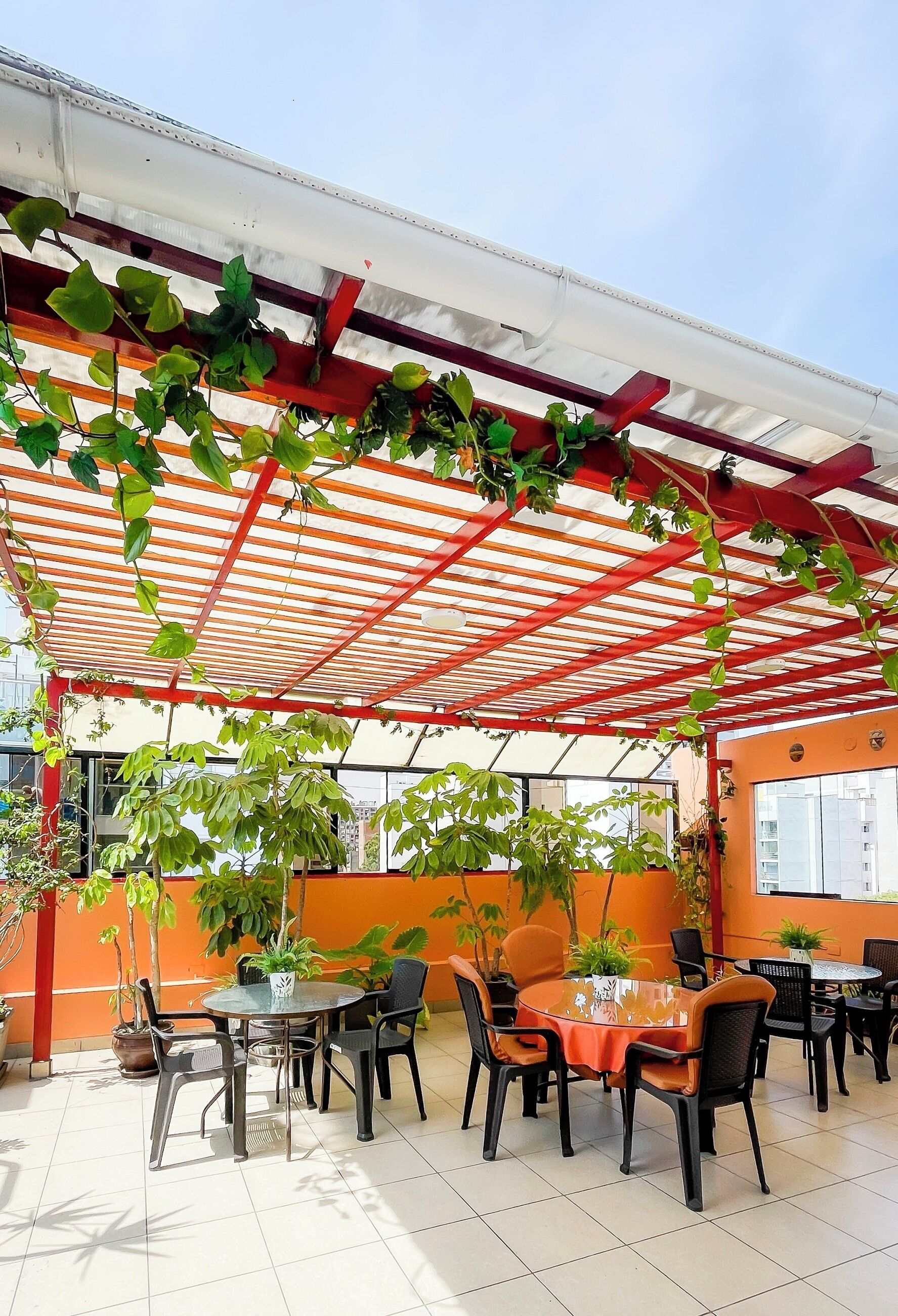 Uma área de pátio ao ar livre com pérgola vermelha, paredes laranja e várias mesas e cadeiras.
