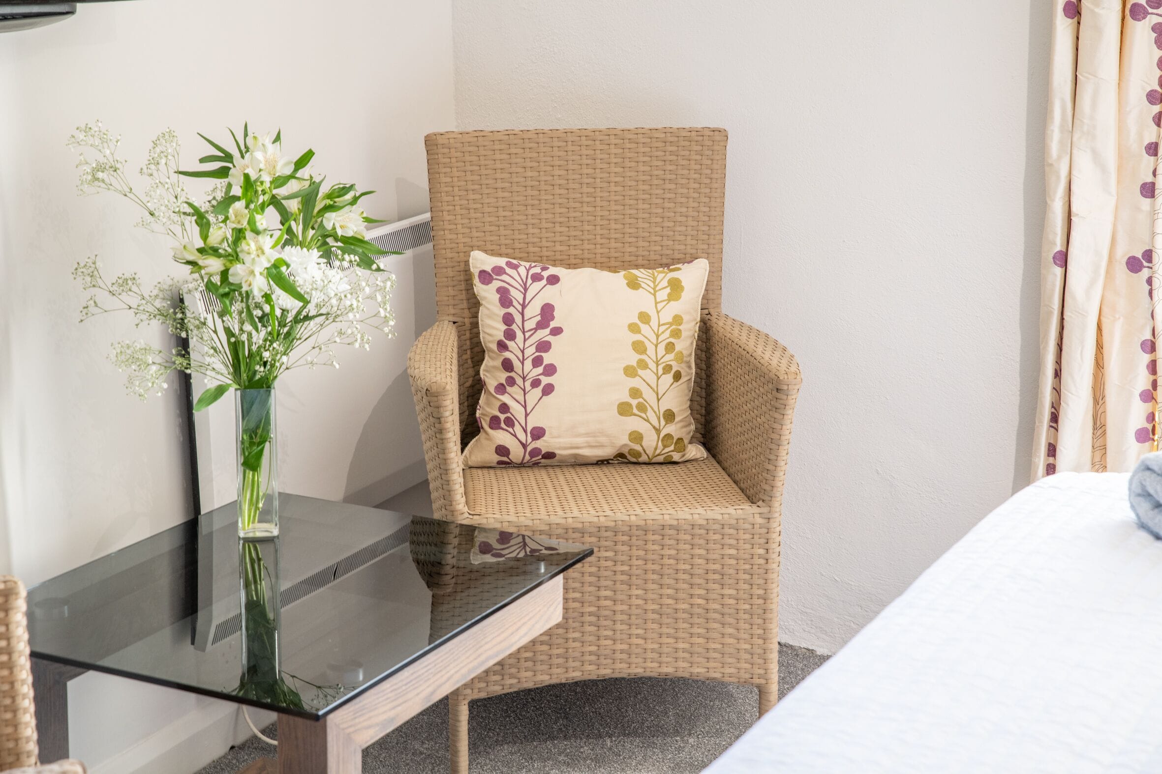 Uma cadeira de vime com almofada, uma mesa de vidro e um vaso com flores.