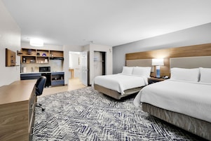 Quarto de hotel com escrivaninha, duas camas, cozinha compacta e tapete estampado.