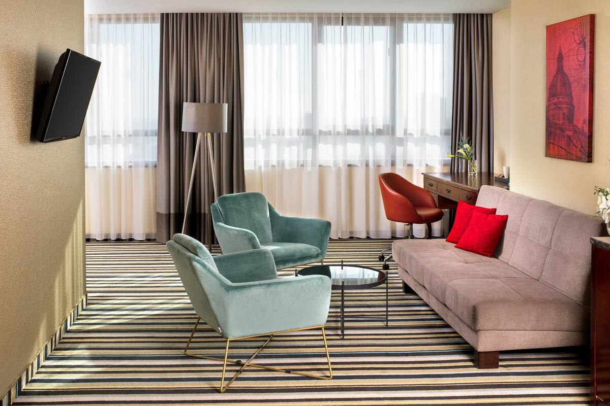 Quarto de hotel com chão listrado, um sofá, duas poltronas, uma TV fixada na parede e um quadro na parede.