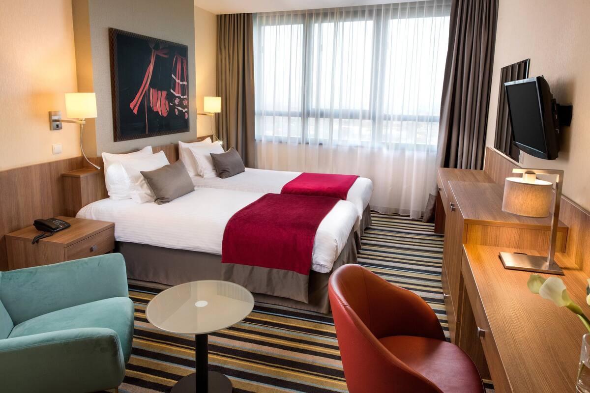 Quarto de hotel com uma cama grande, duas mesas de cabeceira com abajures, uma TV fixada na parede, uma escrivaninha com abajur, uma mesinha e uma cadeira.
