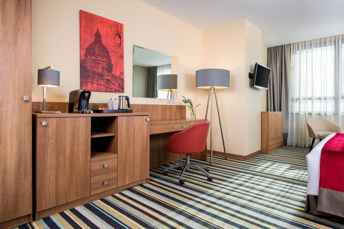 Quarto de hotel com escrivaninha de madeira, uma cadeira vermelha, cama com manta vermelha, televisão e uma janela grande com cortinas.