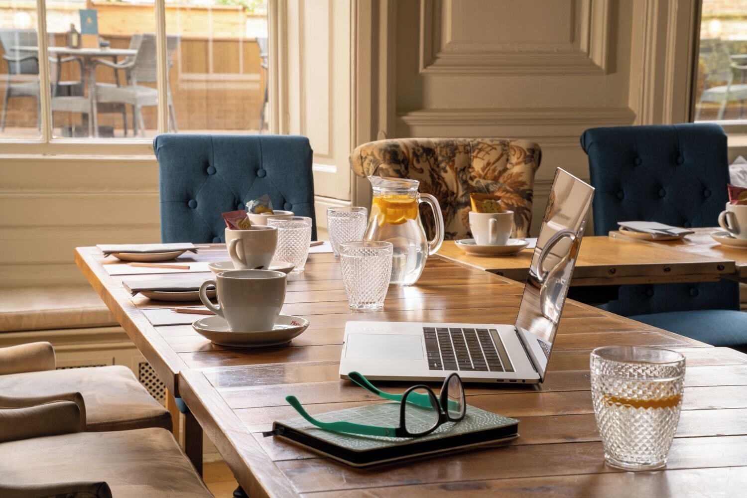 Uma mesa de madeira com um laptop, um caderno, óculos e um jarro de chá.