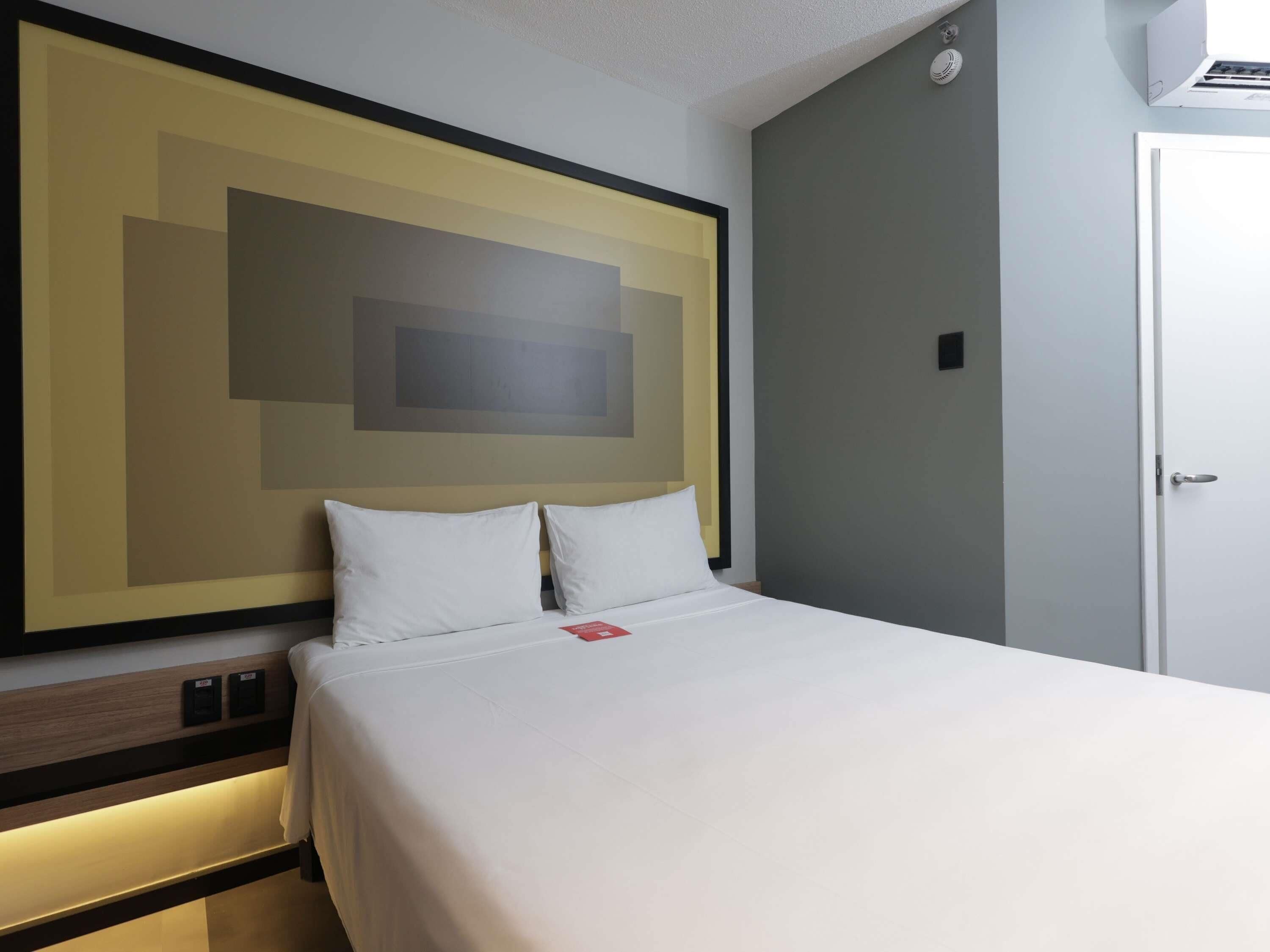 Quarto de hotel com uma cama, um quadro abstrato grande na parede, uma porta e um aparelho de ar condicionado.