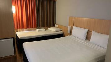 Quarto de hotel com duas camas, um pequeno frigorífico e uma janela com cortinas.