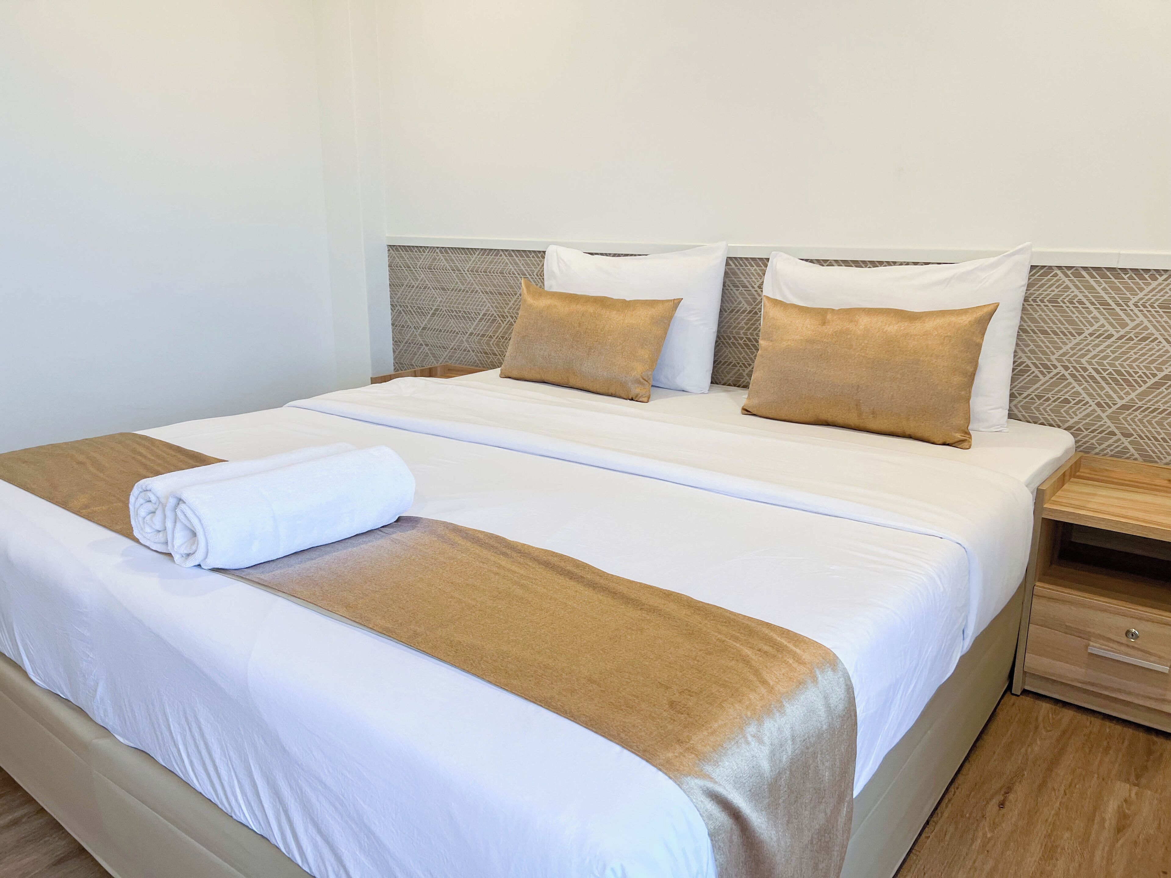 Uma cama bem arrumada com lençóis brancos e duas almofadas castanhas, uma mesa de cabeceira de madeira com gaveta, e uma parede estampada atrás da cama.