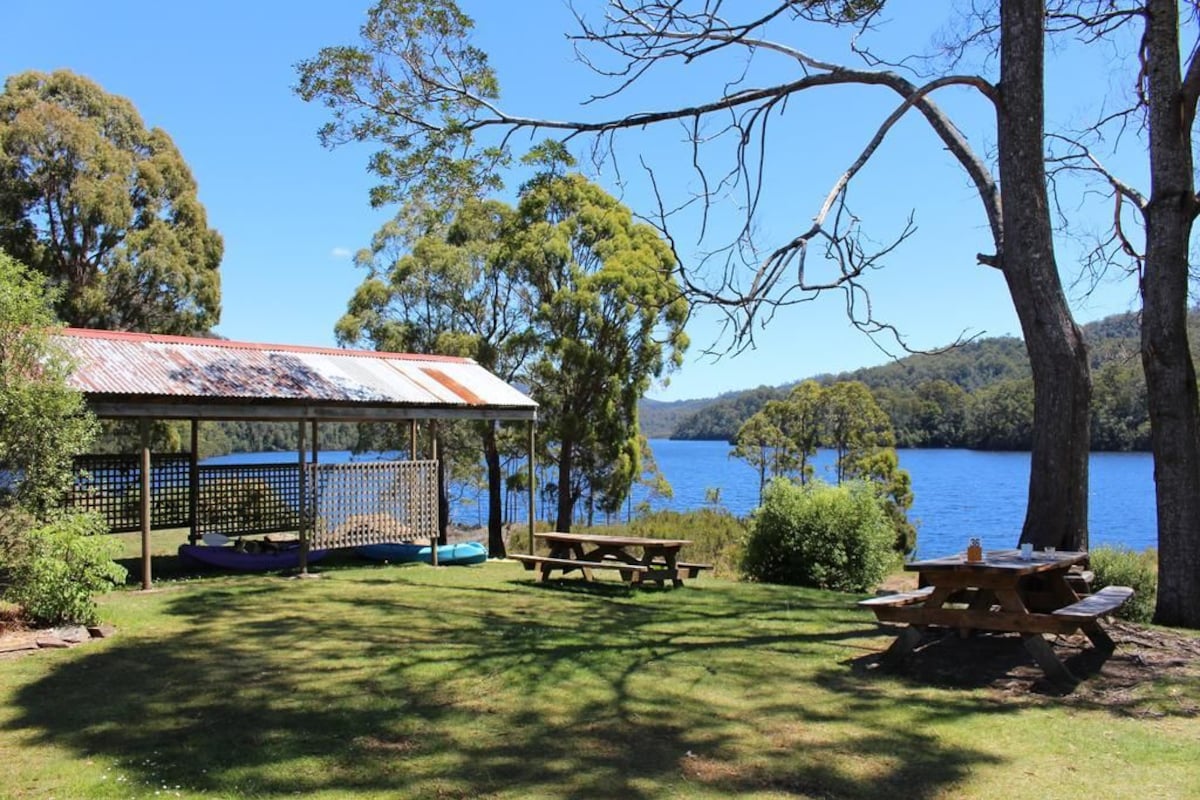 Uma área para piquenique à beira do lago, com um abrigo coberto, mesas de piquenique e vista para o lago.