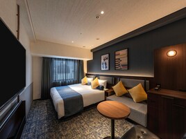 Quarto de hotel com cama, mesas de cabeceira, escrivaninha, cadeira, televisão e janela com cortinas.