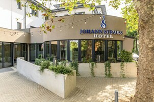 Exterior do Hotel Johann Strauss, com design moderno, apresentando amplas janelas de vidro e área ajardinada com vasos.