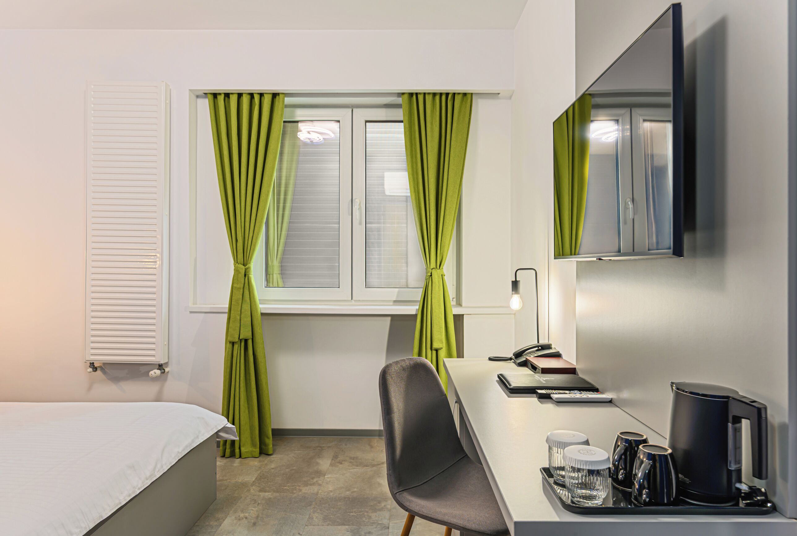 Quarto de hotel com uma cama, uma escrivaninha com cafeteira, uma cadeira e uma janela com cortinas.