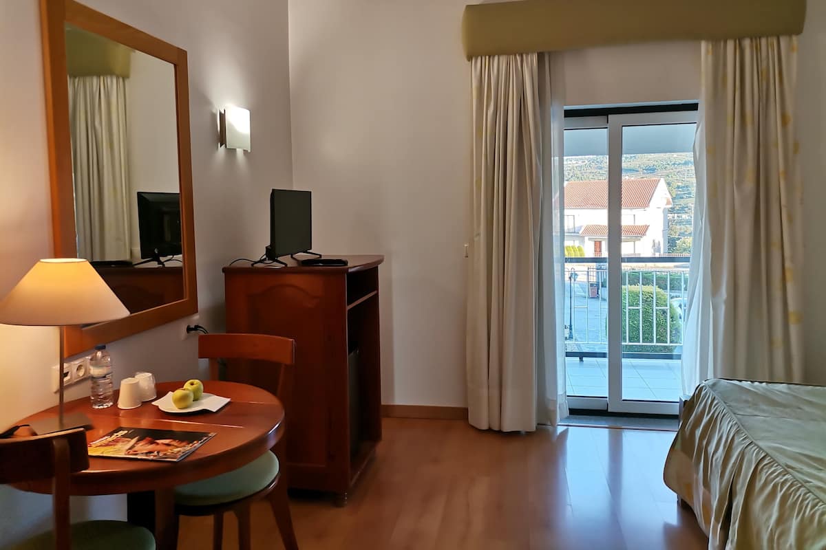 Quarto de hotel com escrivaninha de madeira, cadeira, cama, um espelho grande, televisão sobre um móvel de madeira e vista para um edifício através de uma porta de vidro.