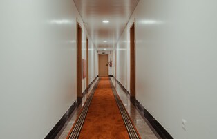 Um corredor comprido e estreito com carpete estampado, paredes brancas e portas de madeira em ambos os lados.