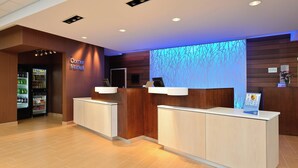 Uma área de recepção de hotel com um balcão de madeira, uma tela digital e um refrigerador repleto de bebidas.