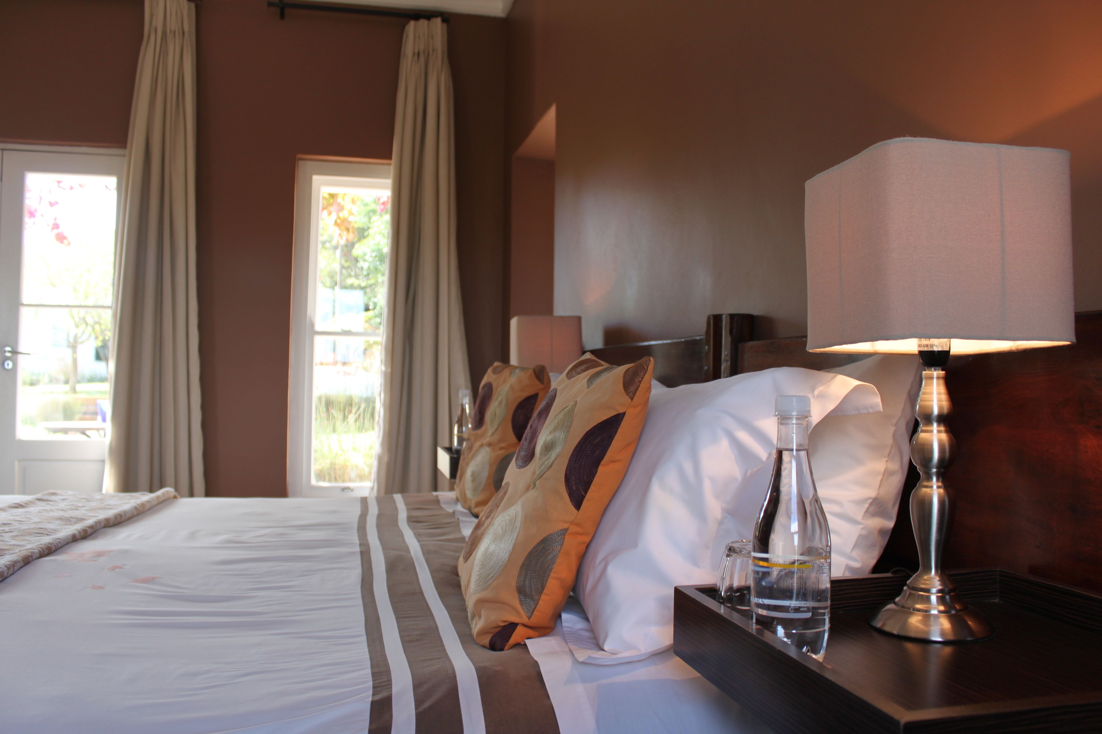 Quarto de hotel com cama, travesseiros, mesa de cabeceira com abajur, garrafa e bandeja.