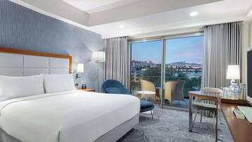 Quarto de hotel com uma cama grande, uma escrivaninha com um computador, uma cadeira e vista da cidade através de uma janela ampla.