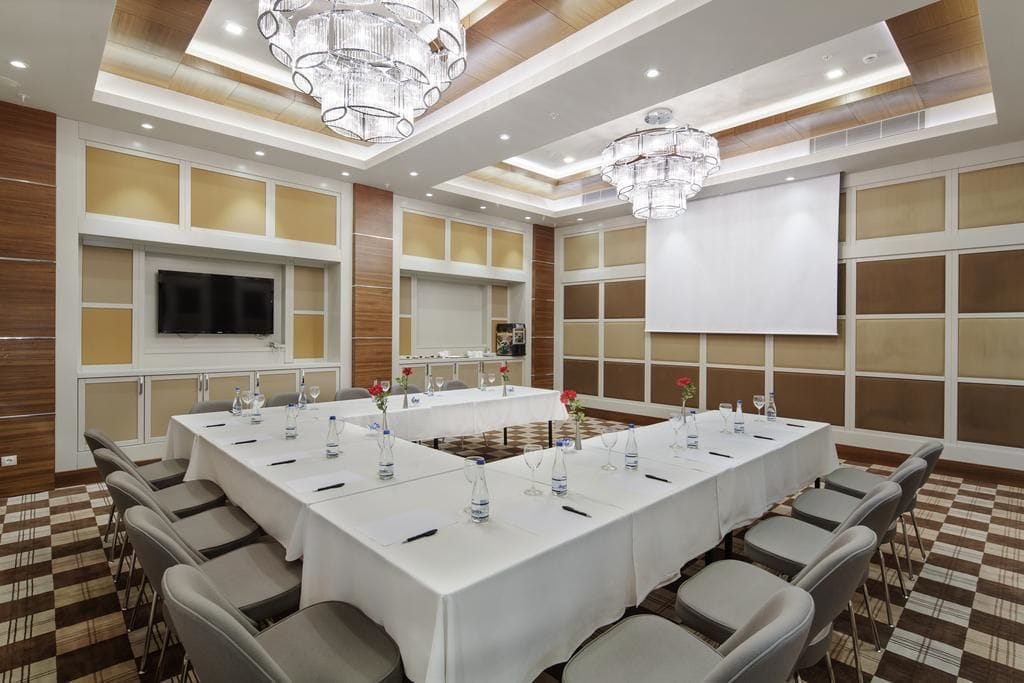 Uma sala de conferências com uma mesa comprida, cadeiras, uma tela de projetor e uma televisão.