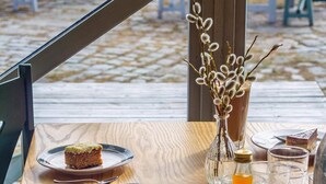 Uma mesa de madeira com uma fatia de bolo, uma vela, um copo de suco de laranja e um vaso com galhos.