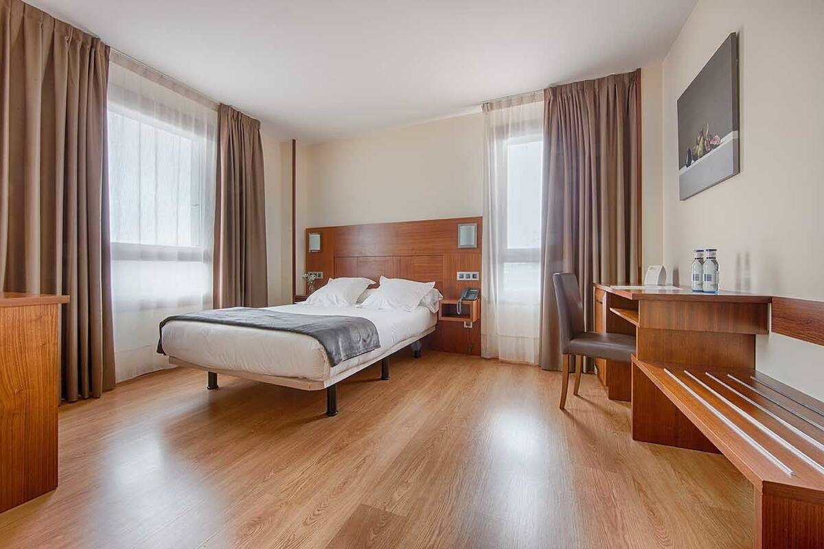 Quarto de hotel com cama de madeira, escrivaninha, cadeira e mesa de cabeceira com telefone.