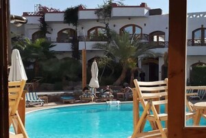 Uma área de piscina com espreguiçadeiras e guarda-sóis, cercada por edifícios com janelas em arco e varandas.