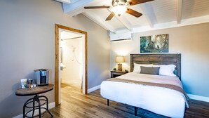 Um quarto com piso de madeira, uma cama com roupa de cama branca, um ventilador de teto, uma mesa de cabeceira com abajur e um quadro na parede.