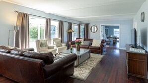 Uma sala de estar ampla com um sofá de couro marrom, uma poltrona branca e um sofá marrom. Há uma mesa de centro com um vaso de flores e uma televisão à direita. O quarto possui janelas grandes com cortinas, uma área de jantar ao fundo e um armário de madeira com a etiqueta "FARMHOUSE".
