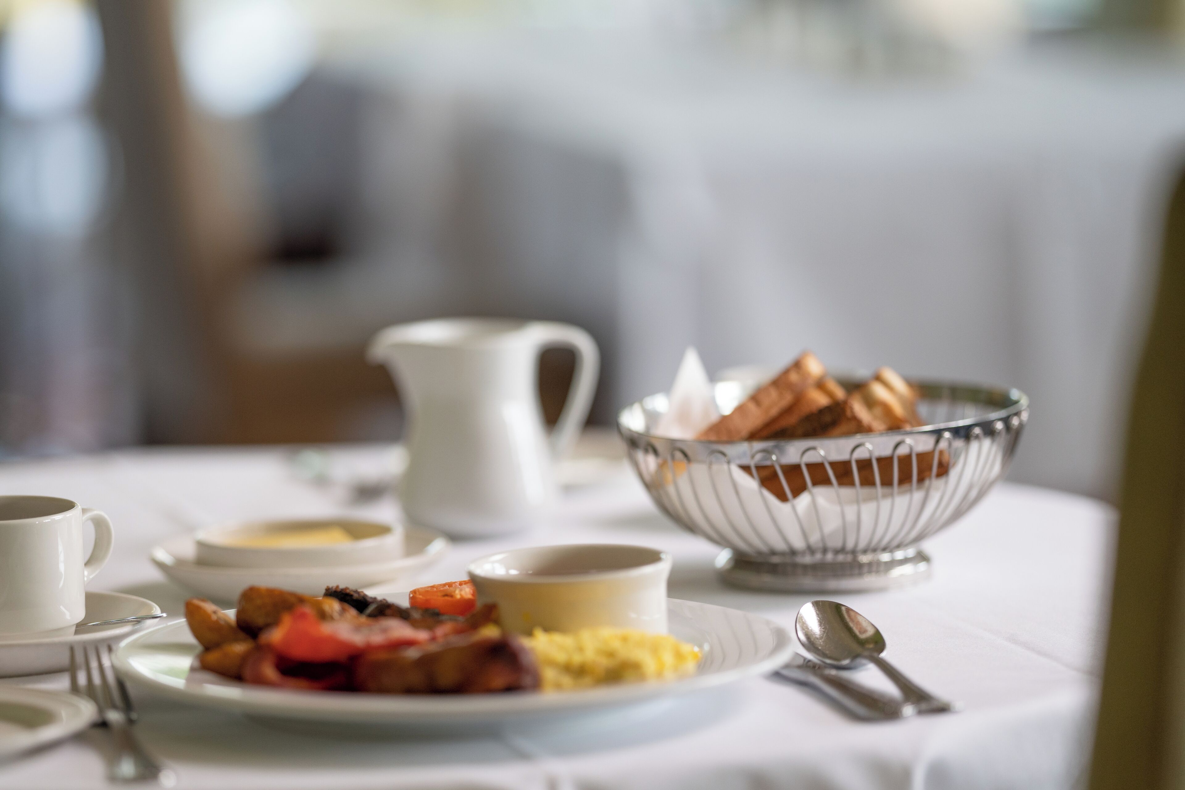 Uma mesa de café da manhã com pratos de comida, uma cesta de torradas, um jarro e uma xícara.