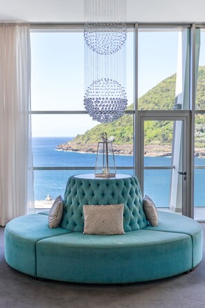Um sofá azul-esverdeado com encosto estofado e duas almofadas, posicionado em frente a uma grande janela com vista para o mar e uma paisagem montanhosa.